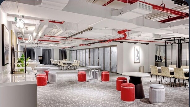 Um espaço de escritório moderno, com paredes brancas, detalhes em vermelho e uma variedade de opções de assento, incluindo mesas e cadeiras.