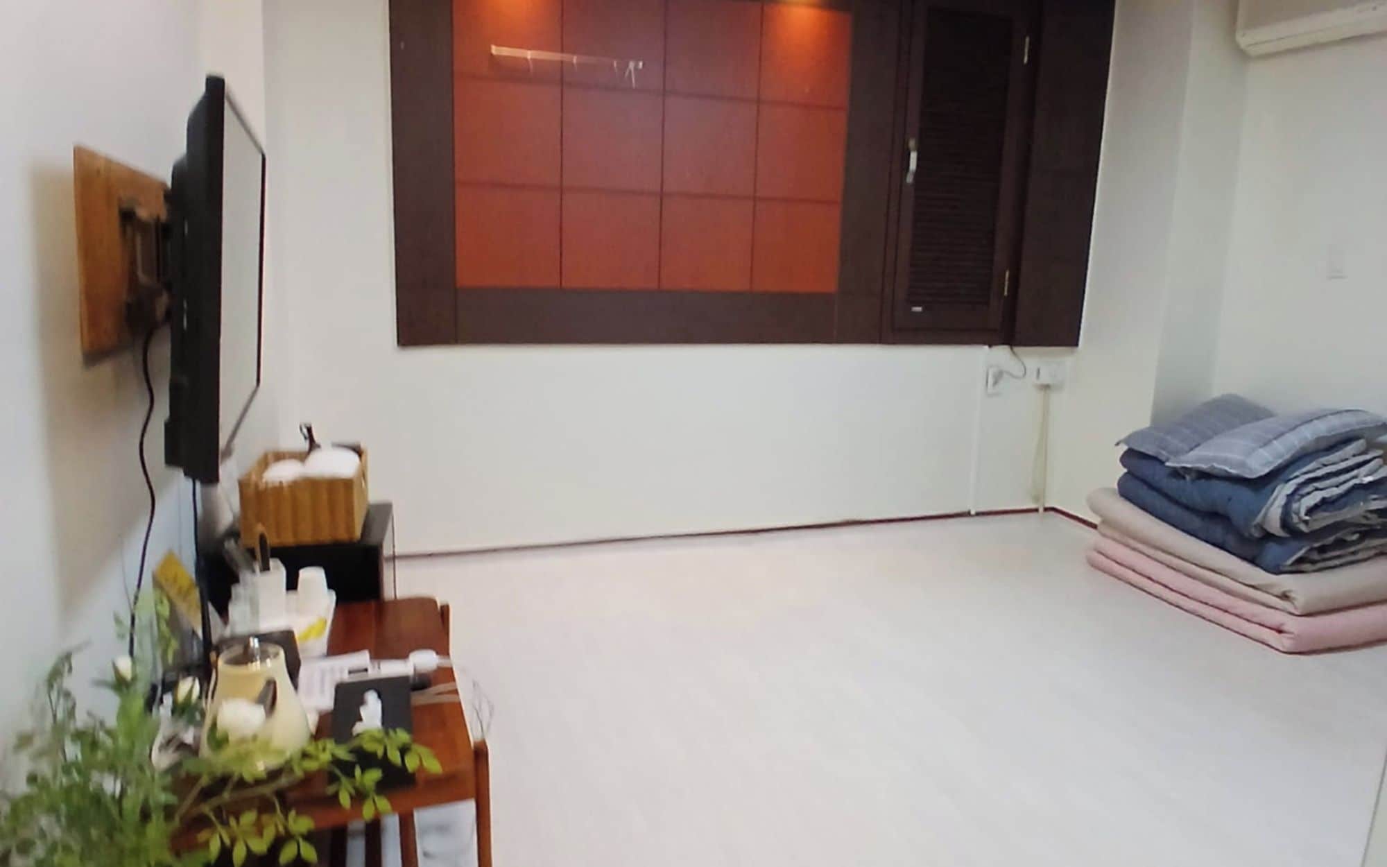 Quarto com uma mesa de madeira, televisão presa na parede, janela com persianas e uma pilha de roupas dobradas.