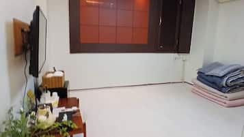 Quarto com uma mesa de madeira, televisão presa na parede, janela com persianas e uma pilha de roupas dobradas.