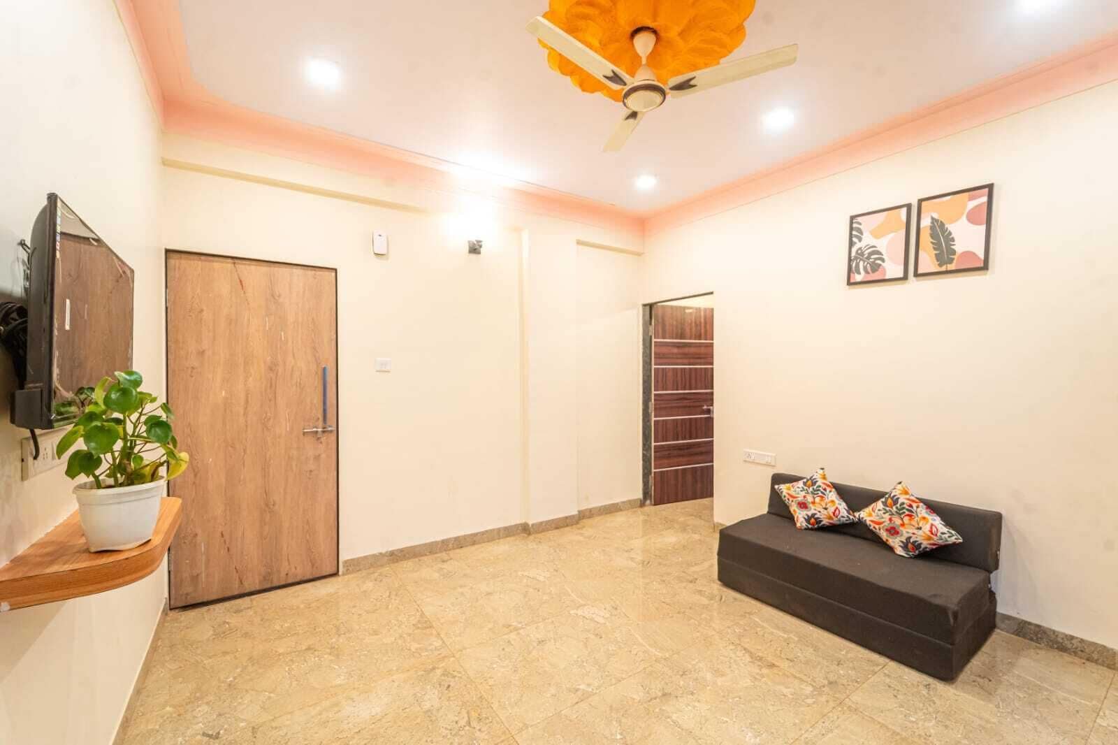 Um quarto com ventilador de teto, sofá com almofadas estampadas, planta em vaso e porta de madeira.