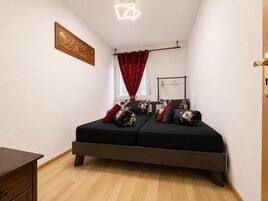 Quarto com uma cama, cômoda de madeira, janela com cortinas vermelhas e luminária fixada na parede.