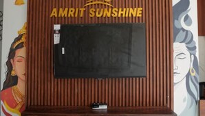 Uma televisão fixada na parede, com design em painel de madeira, um relógio acima e o texto 'AMRIT SUNSHINE' abaixo.