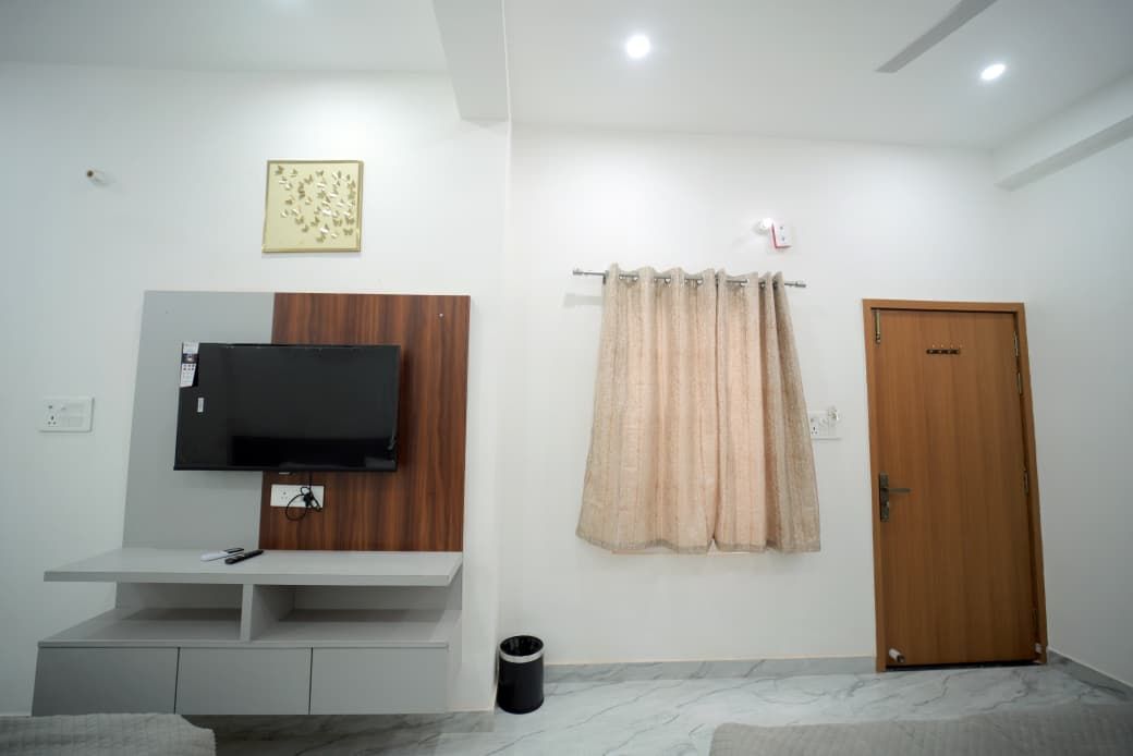 Quarto com televisão de tela plana presa a um painel de madeira, barra de cortina com cortina bege, porta de madeira e lixeira.
