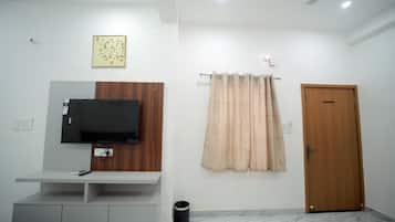 Quarto com televisão de tela plana presa a um painel de madeira, barra de cortina com cortina bege, porta de madeira e lixeira.