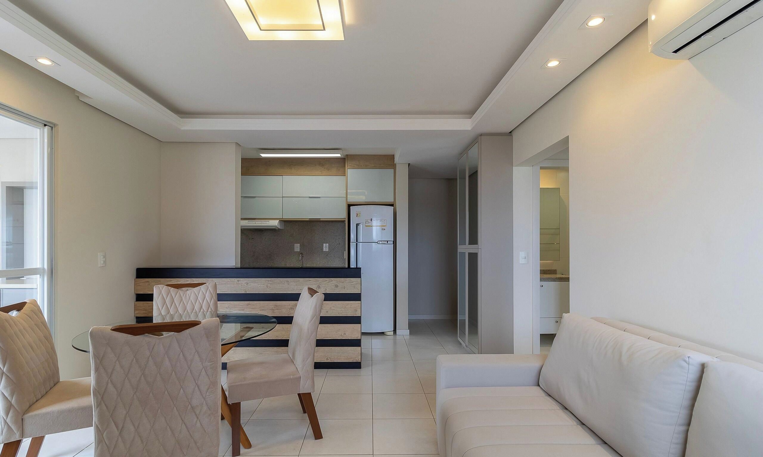 Uma sala de estar moderna com sofá e poltronas, uma área de jantar com mesa e cadeiras, e uma cozinha com geladeira e armários.