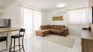 Uma sala de estar moderna com sofá seccional, uma mesa de jantar com cadeiras, área de cozinha com micro-ondas e uma grande janela com cortinas.