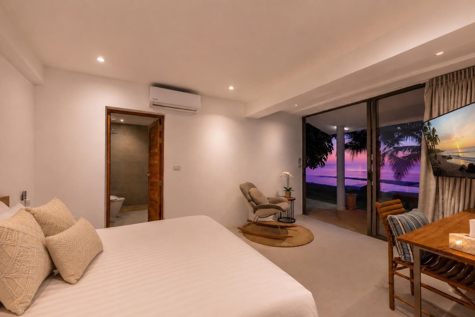 Quarto de hotel com cama, poltrona, mesa e televisão exibindo uma praia.