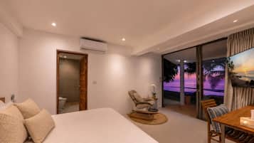 Quarto de hotel com cama, poltrona, mesa e televisão exibindo uma praia.