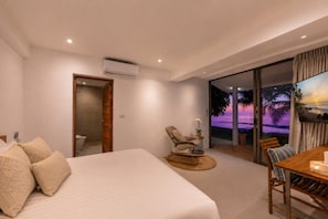 Quarto de hotel com cama, poltrona, mesa e televisão exibindo uma praia.