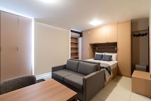 Quarto compacto com cama, sofá, pequena mesa e espaço de armazenamento embutido.
