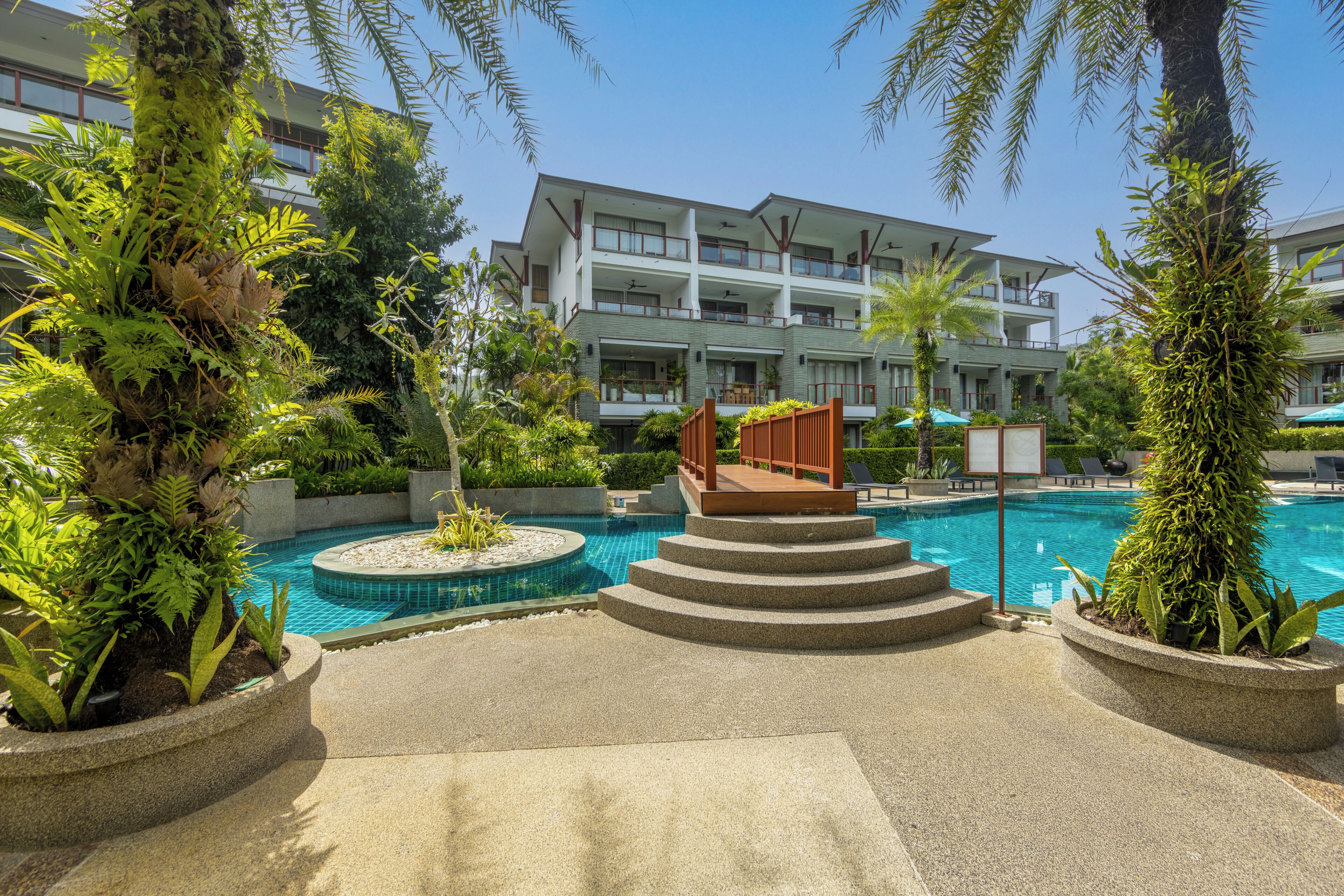 Uma área de piscina com um elemento aquático circular, degraus que levam a um deck de madeira e um edifício de vários andares ao fundo.
