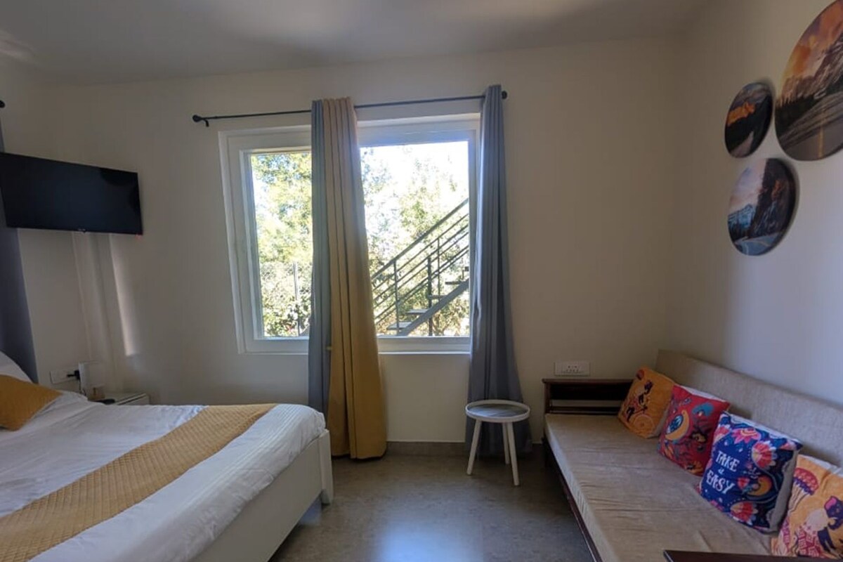 Quarto com duas camas, janela com cortinas, um banquinho pequeno e decoração circular na parede.