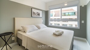 Quarto moderno com uma cama grande, uma mesa pequena, um quadro emoldurado e uma janela com vista para prédios.