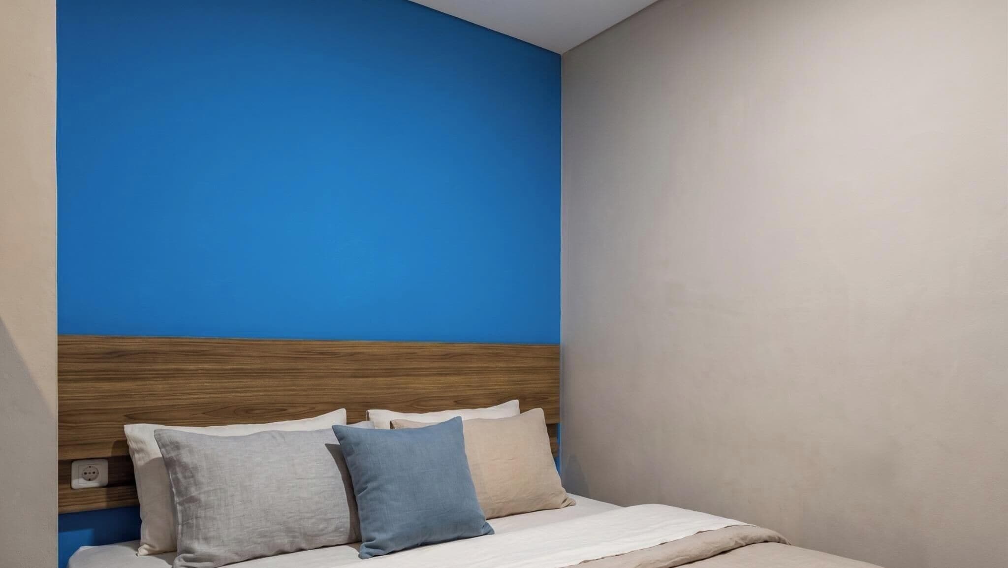 Quarto com parede azul de destaque, cabeceira de madeira e cama com várias almofadas.
