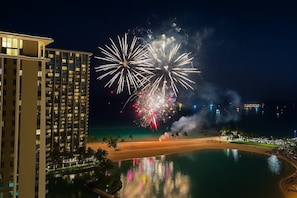 Hotel na orla marítima com piscina, praia de areia e vista para fogos de artifício ao longe.