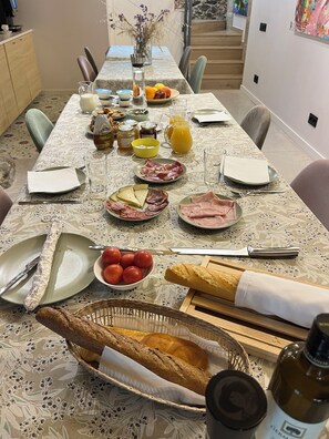 Uma mesa de jantar posta com diversos alimentos, incluindo pão, tomates e carnes, em um ambiente com escada e um vaso de flores.