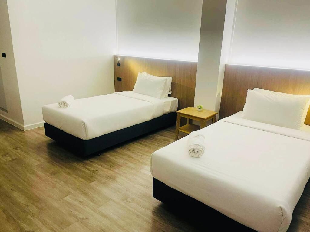 Quarto de hotel com duas camas de solteiro, uma pequena mesa de cabeceira de madeira e luminária fixada na parede.
