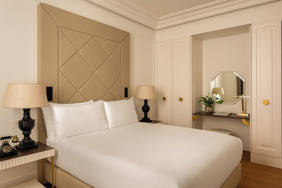Quarto de hotel com uma cama grande, duas mesas de cabeceira, um criado-mudo, um espelho e uma pequena prateleira com uma planta.