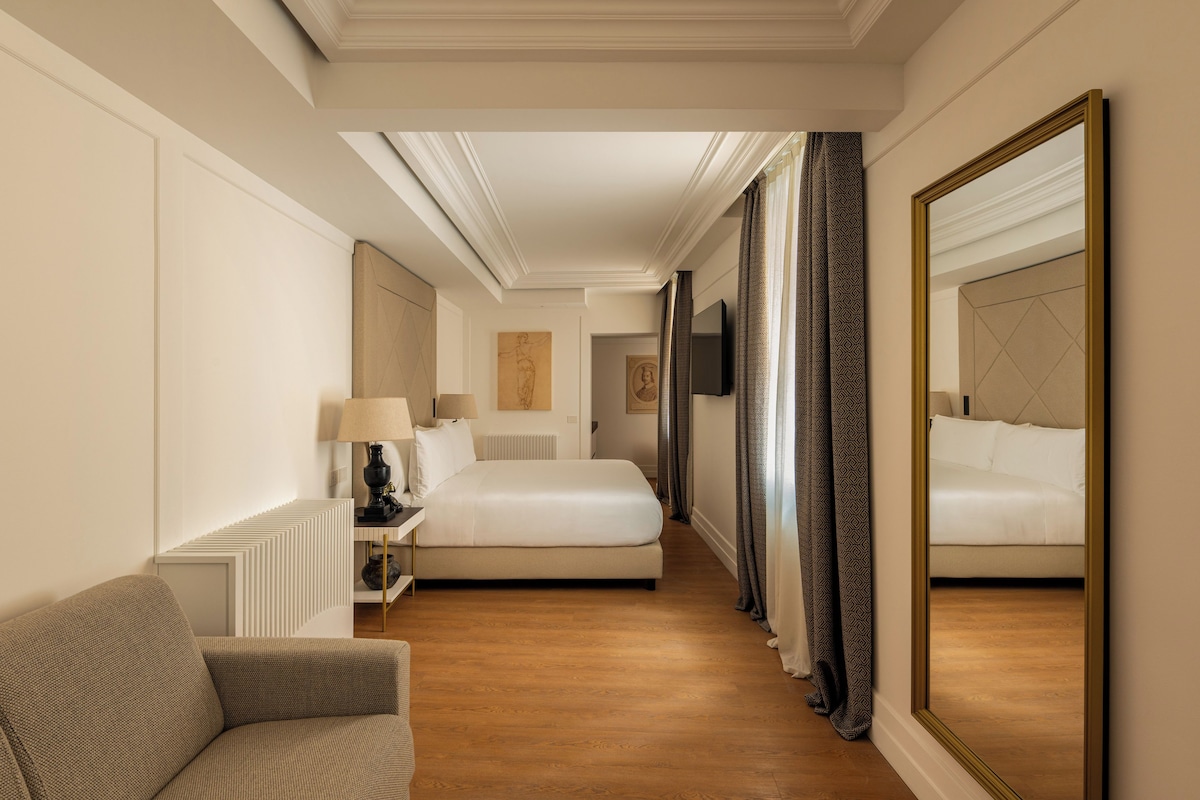 Quarto de hotel com uma cama grande, área de estar com uma poltrona, escrivaninha e um espelho amplo.