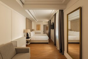 Quarto de hotel com uma cama grande, área de estar com uma poltrona, escrivaninha e um espelho amplo.