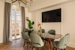 Uma sala de jantar com mesa de vidro, quatro cadeiras, um vaso com flores, uma TV de tela plana grande na parede e vista para o exterior através de uma porta de vidro.
