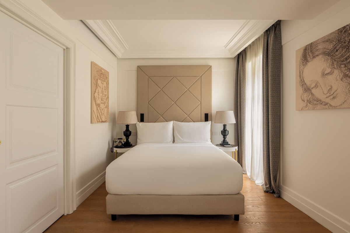 Quarto de hotel com uma cama grande, duas mesas de cabeceira com luminárias, cabeceira com padrão geométrico, uma porta e uma parede com uma obra de arte emoldurada.