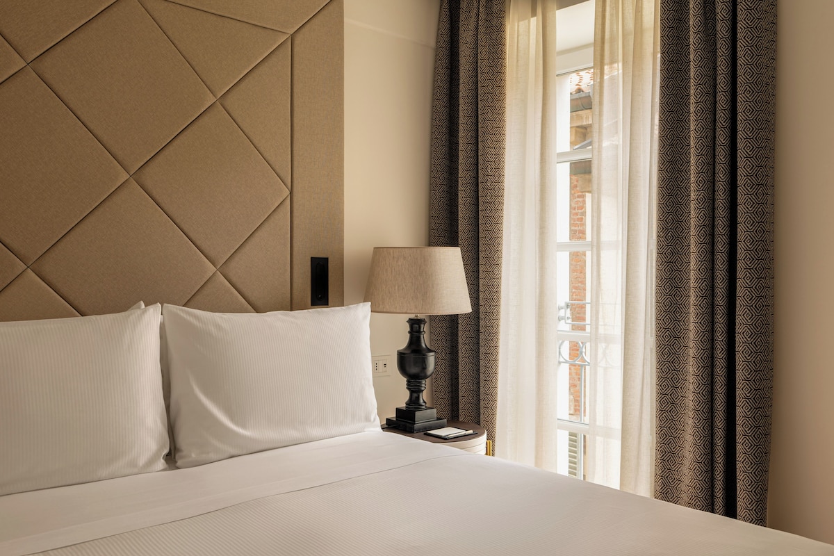 Quarto de hotel com cama, mesa de cabeceira, luminária e janela com cortinas.