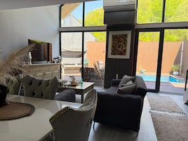 Uma sala moderna com um sofá, uma mesa de jantar e uma televisão. Há uma grande janela com vista para um quintal com piscina.