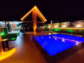 Piscina com borda iluminada em azul, deck de madeira, área de estar externa e um edifício com telhado triangular iluminado por luzes quentes.