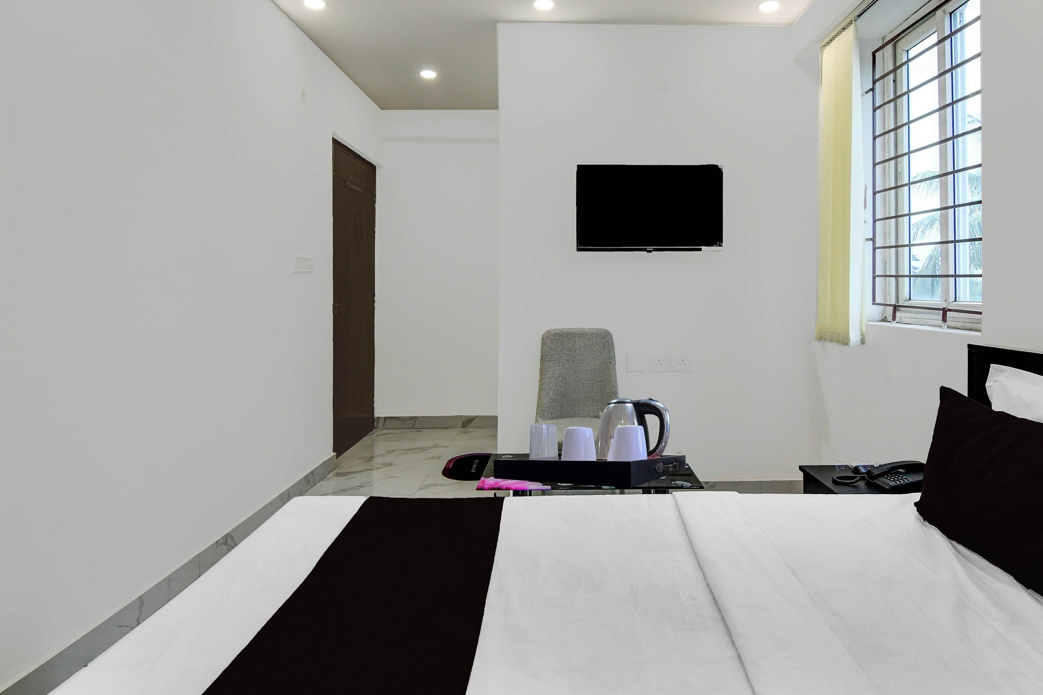 Quarto de hotel com uma cama, televisão de tela plana na parede, janela com persianas e uma pequena mesa com chaleira e xícaras.