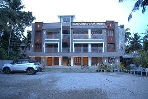 Um edifício de vários andares com a inscrição 'NAKSHATRA APARTMENTS' na fachada, com varandas, entrada protegida por portão e área de estacionamento com um carro e uma bicicleta.