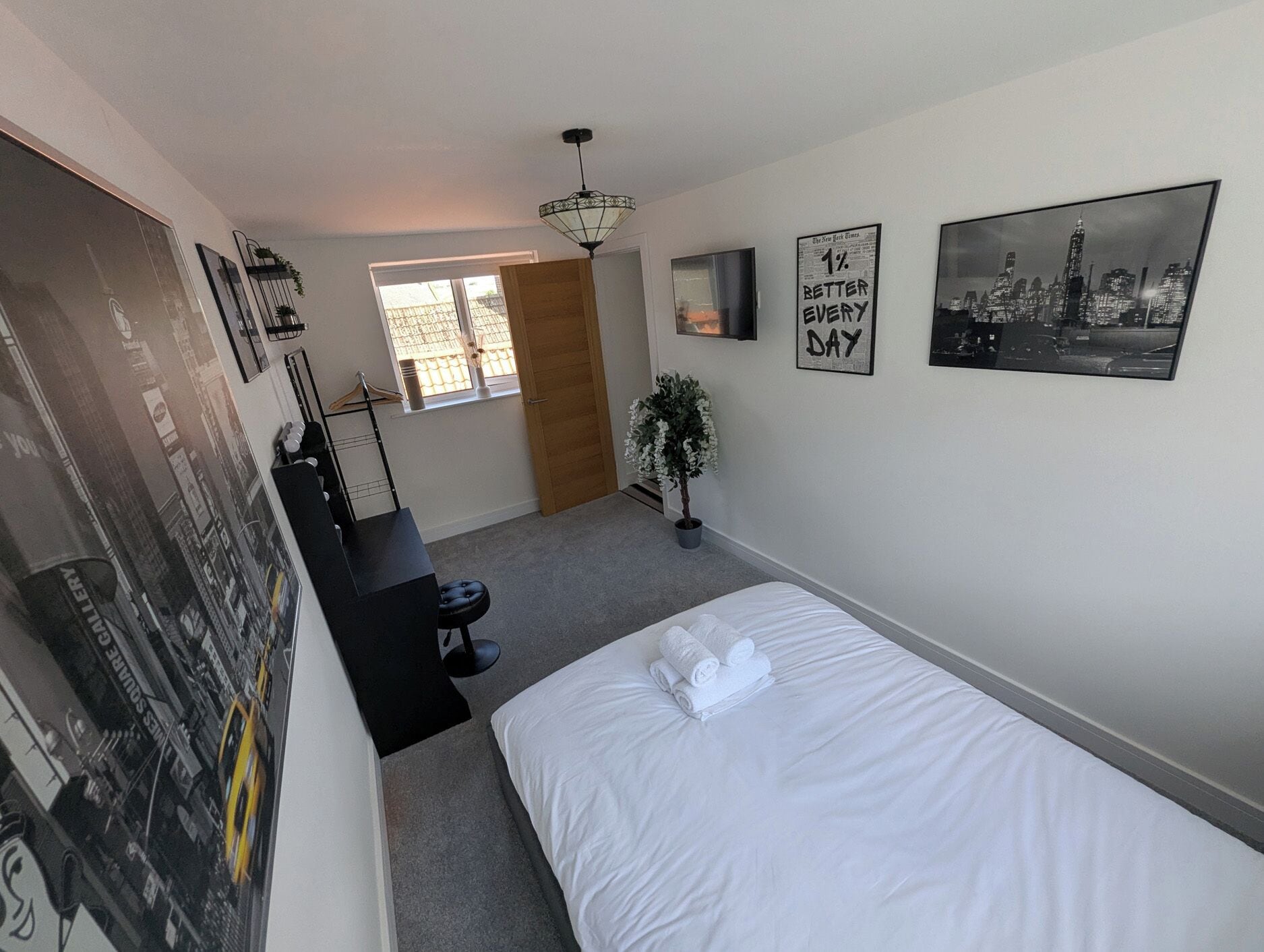 Quarto de hotel com cama, escrivaninha, cadeira, planta pendente, um pôster e janela com vista para a cidade.