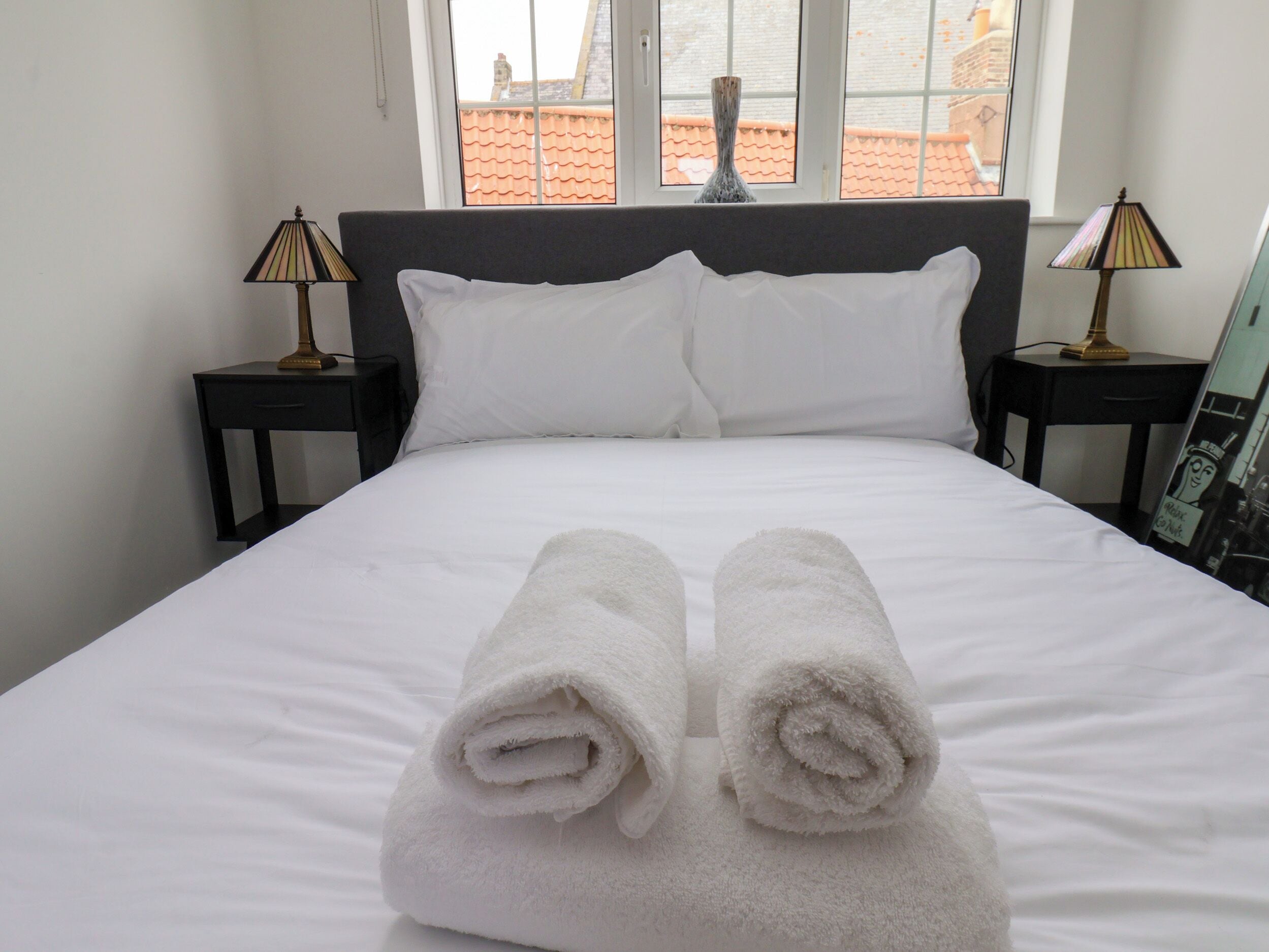 Cama bem arrumada com lençóis brancos e duas toalhas enroladas no centro, ladeada por duas mesas de cabeceira com abajures, em um quarto com janela para um telhado de telhas vermelhas.