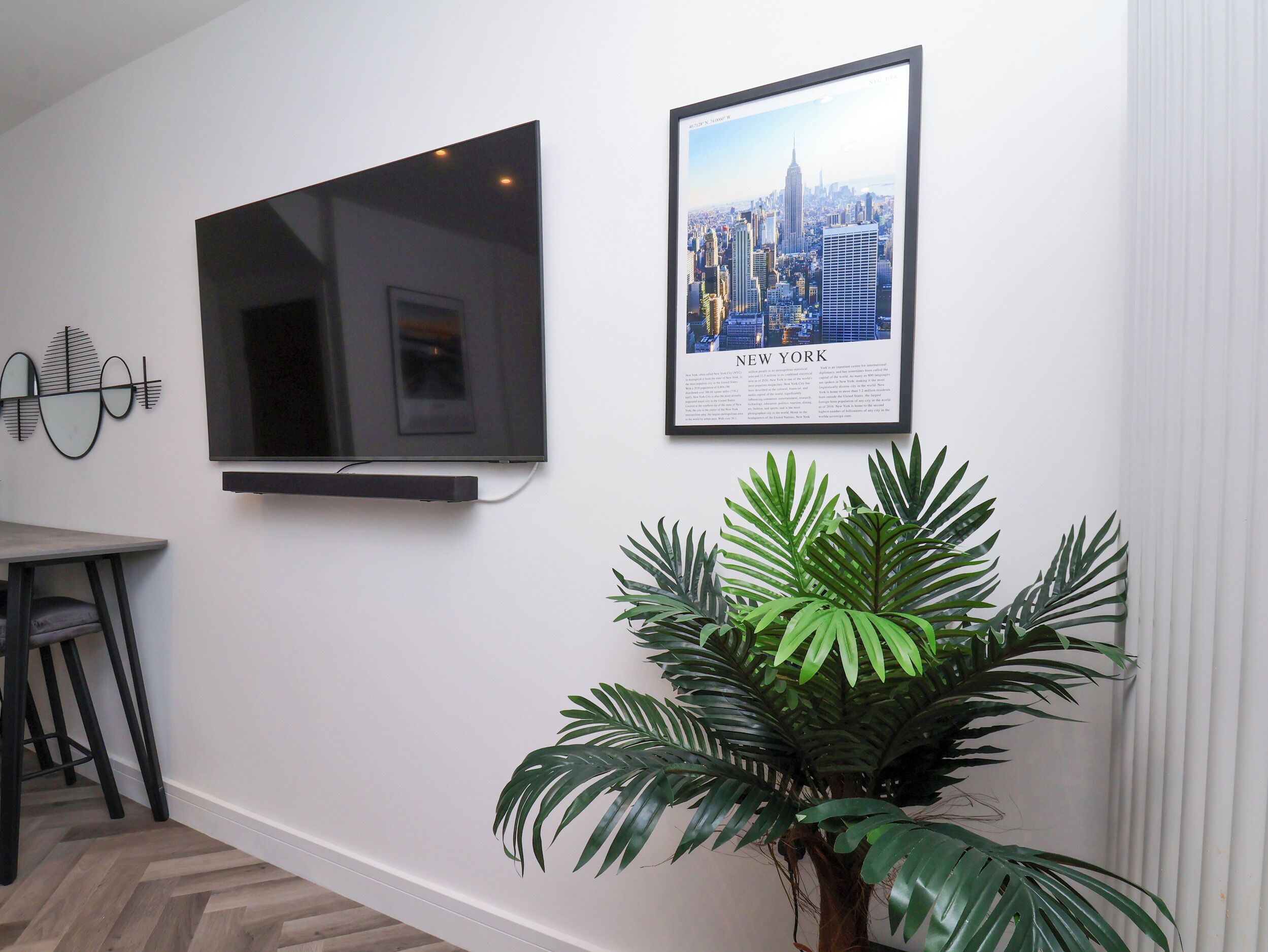 Quarto moderno com televisão de tela plana na parede, um cartaz com vista da cidade emoldurada e um vaso de planta.