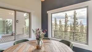 Uma área de jantar moderna com uma mesa redonda de madeira, um vaso com flores e grandes janelas com vista para árvores e montanhas.