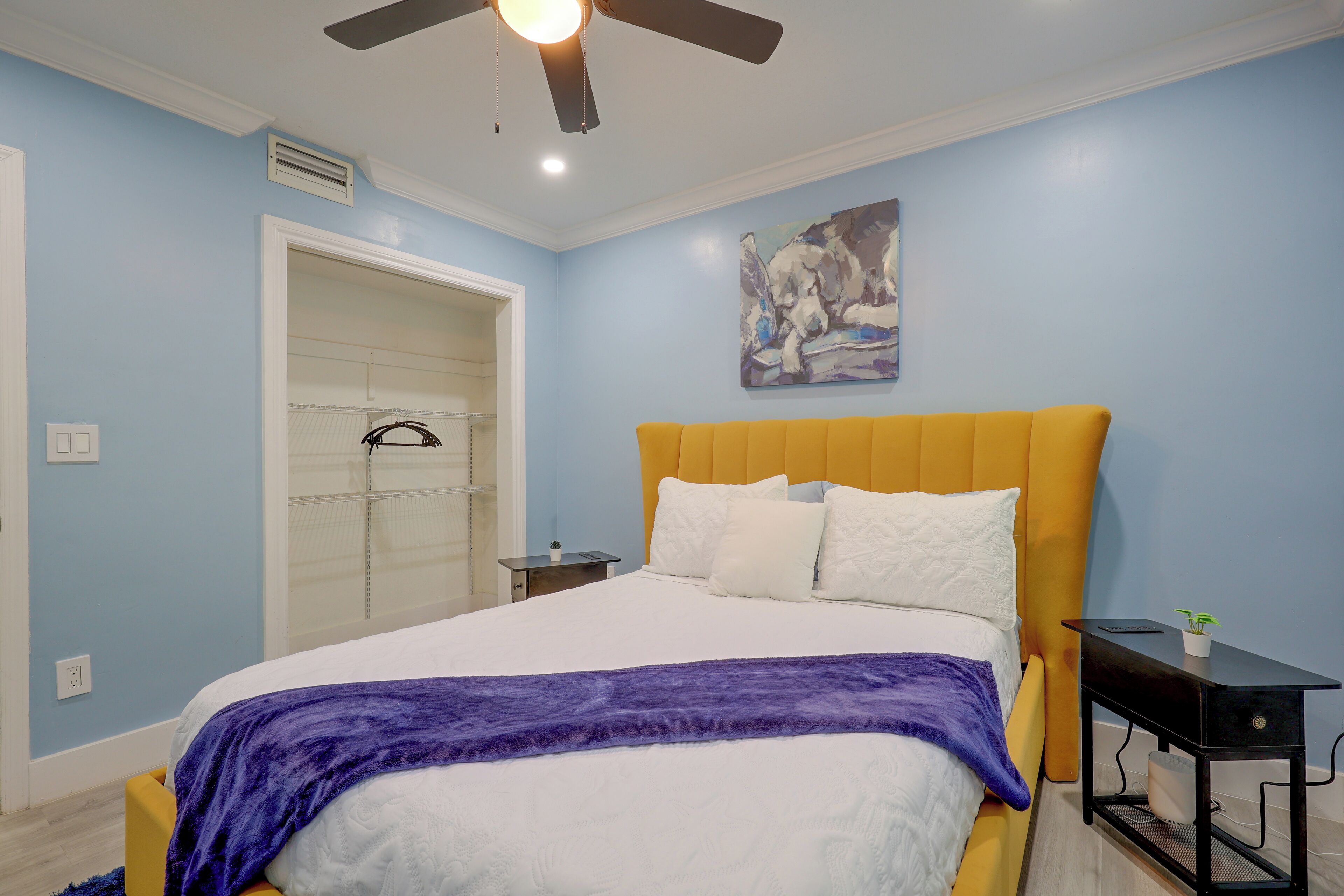 Quarto com cama, mesa de cabeceira, ventilador de teto, pintura e porta com armário.