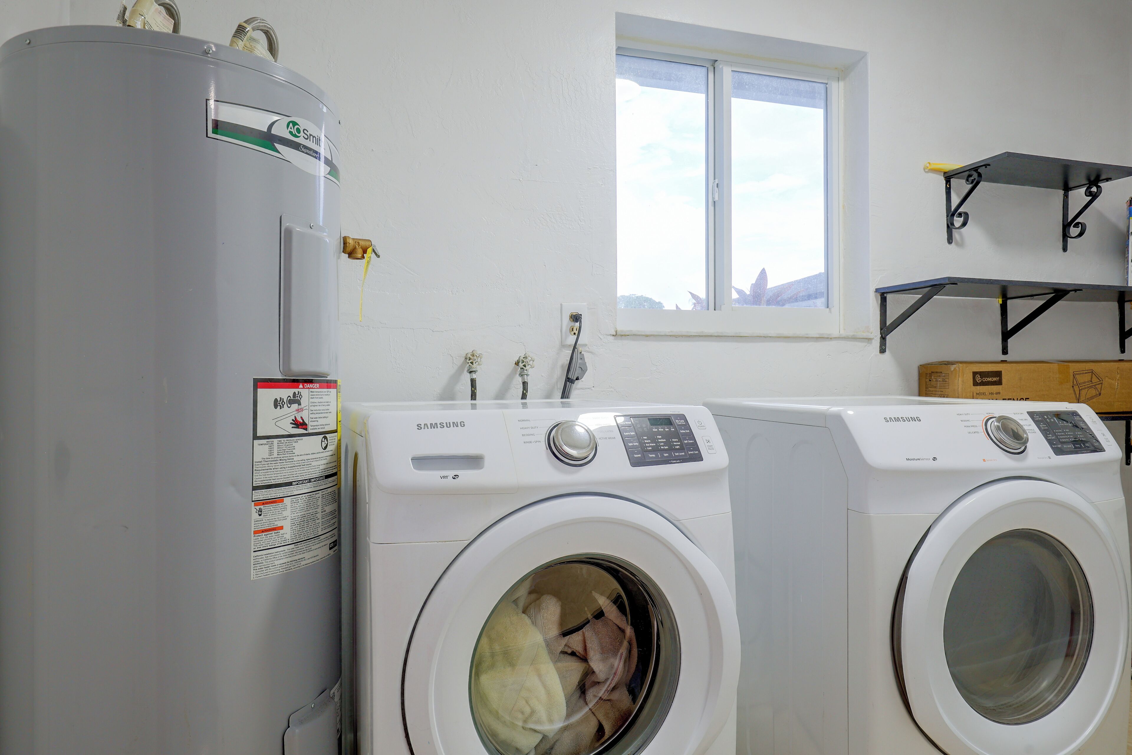 A laundry area with two white washing machines, a water heater, and a window with a view.