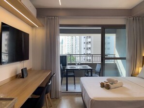 Quarto de hotel com cama, secretária com cadeira, televisão e varanda com mesa e cadeiras.