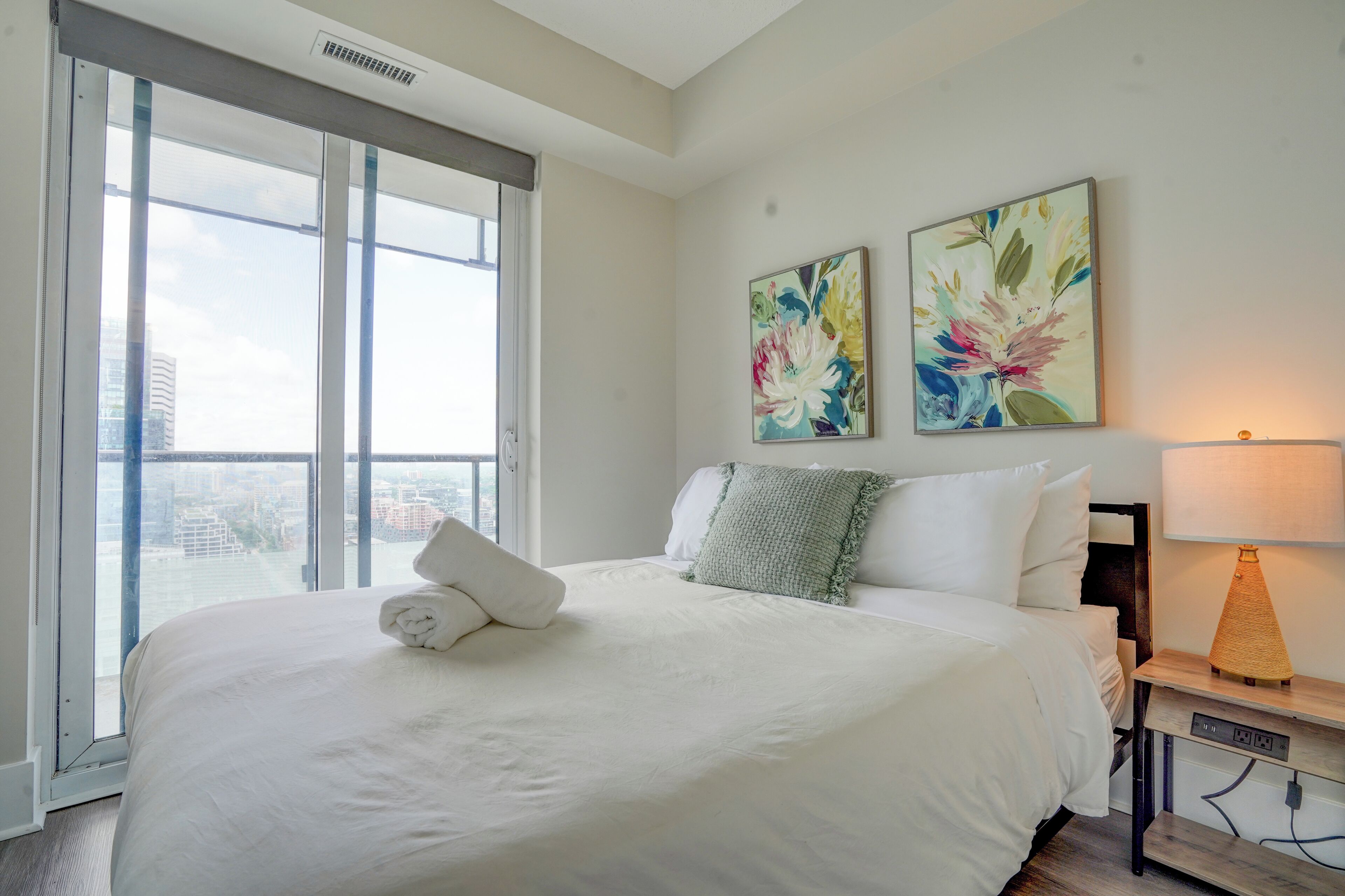 Quarto de hotel com uma cama grande, uma janela com vista para a cidade e duas pinturas florais na parede.