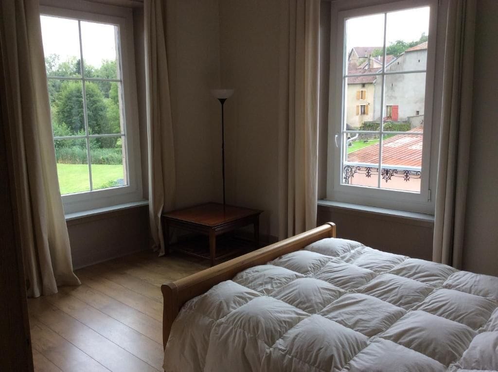 Quarto com cama, duas janelas, luminária de chão e vista para um jardim e edifícios.
