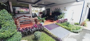 Uma área de recepção com um banco de madeira, plantas em vasos e uma rampa que dá acesso à entrada.