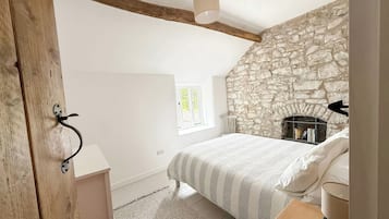 Quarto com lareira de pedra, cama com colcha estampada, vigas de madeira no teto e uma pequena mesa com abajur.