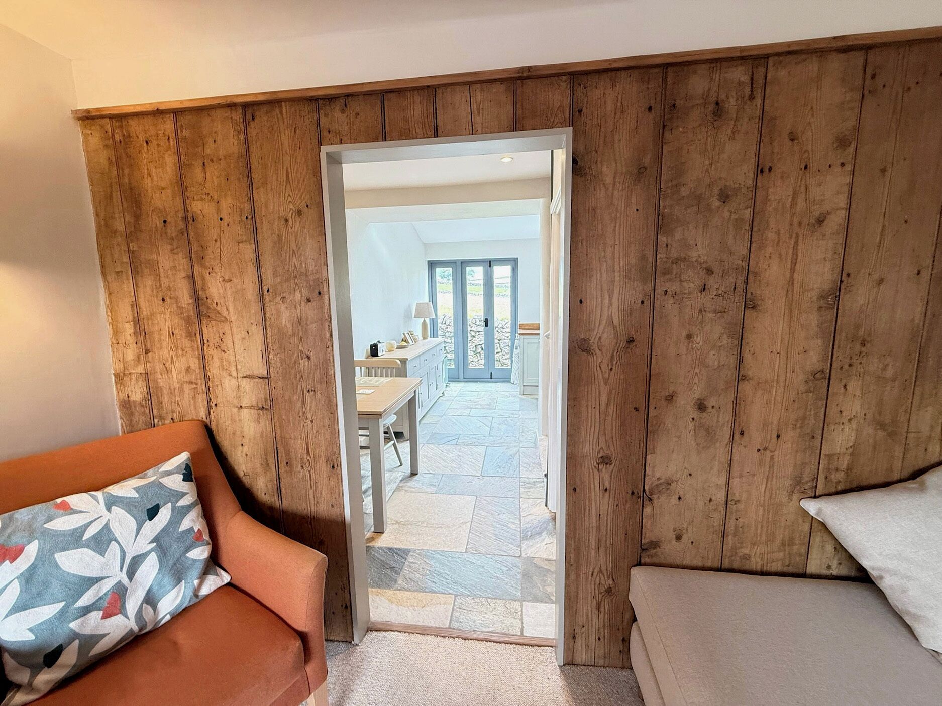 Um quarto com parede de madeira, um sofá, uma cadeira e uma porta que dá para uma área de jantar.
