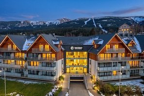 Um hotel com vários andares, entrada central e varandas, rodeado por montanhas cobertas de neve.