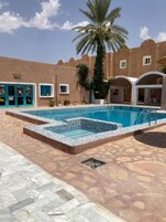 Uma piscina com deck revestido, uma palmeira e um edifício com janelas em arco.