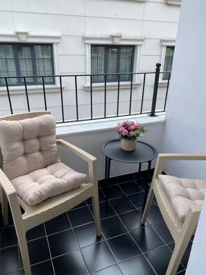 Uma varanda com duas cadeiras de vime, uma mesa redonda pequena com um vaso de planta e piso de azulejos.
