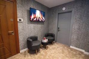 Um quarto com porta de madeira, parede cinzenta, televisão mostrando uma lareira e dois poltronas cinzas com uma pequena mesa no meio.