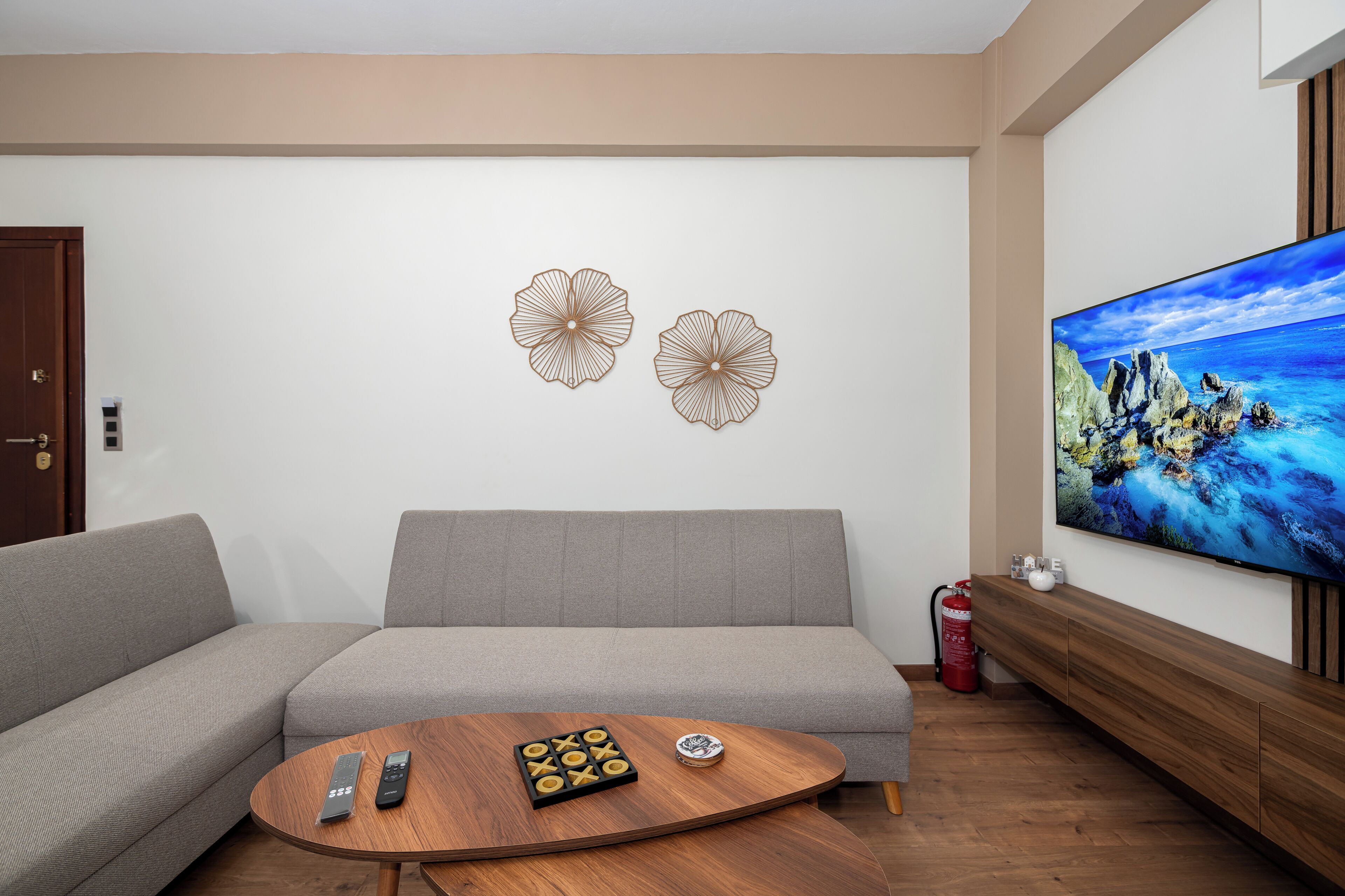 Uma sala de estar moderna com um sofá cinzento, uma mesa de centro de madeira com controles remotos e uma televisão grande exibindo uma paisagem cênica.