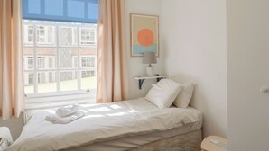 Quarto com uma cama, uma mesinha de cabeceira com abajur, uma janela com cortinas e uma prateleira na parede com um quadro.