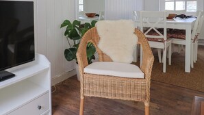 Um quarto com televisão sobre um móvel branco, uma cadeira de vime com almofada branca, uma mesa de jantar com cadeiras e uma planta pendente.